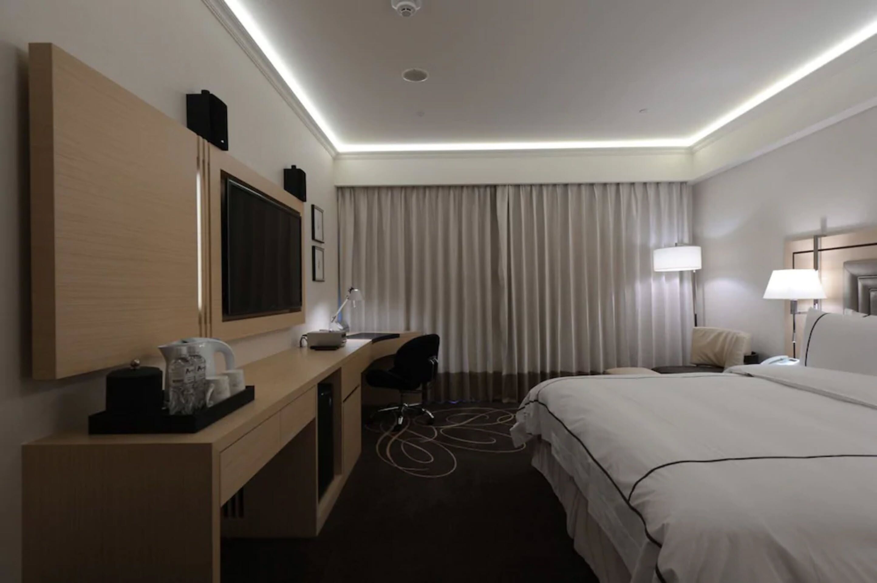 Quarto de hotel com cama, escrivaninha com cadeira, televisão, luminária e janela com cortinas.