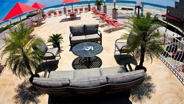 Um espaço de lazer à beira-mar, com espreguiçadeiras, uma mesa redonda e guarda-sóis vermelhos.
