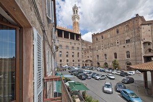 Pátio com um edifício grande, com janelas em arco e uma torre. Estacionamento com vários carros.