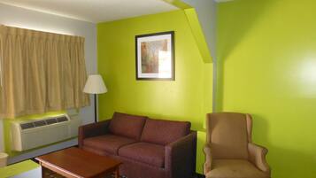 Quarto com parede verde, sofá castanho, mesa de madeira, poltrona bege, quadro emoldurado, janela com cortinas bege e piso xadrez.