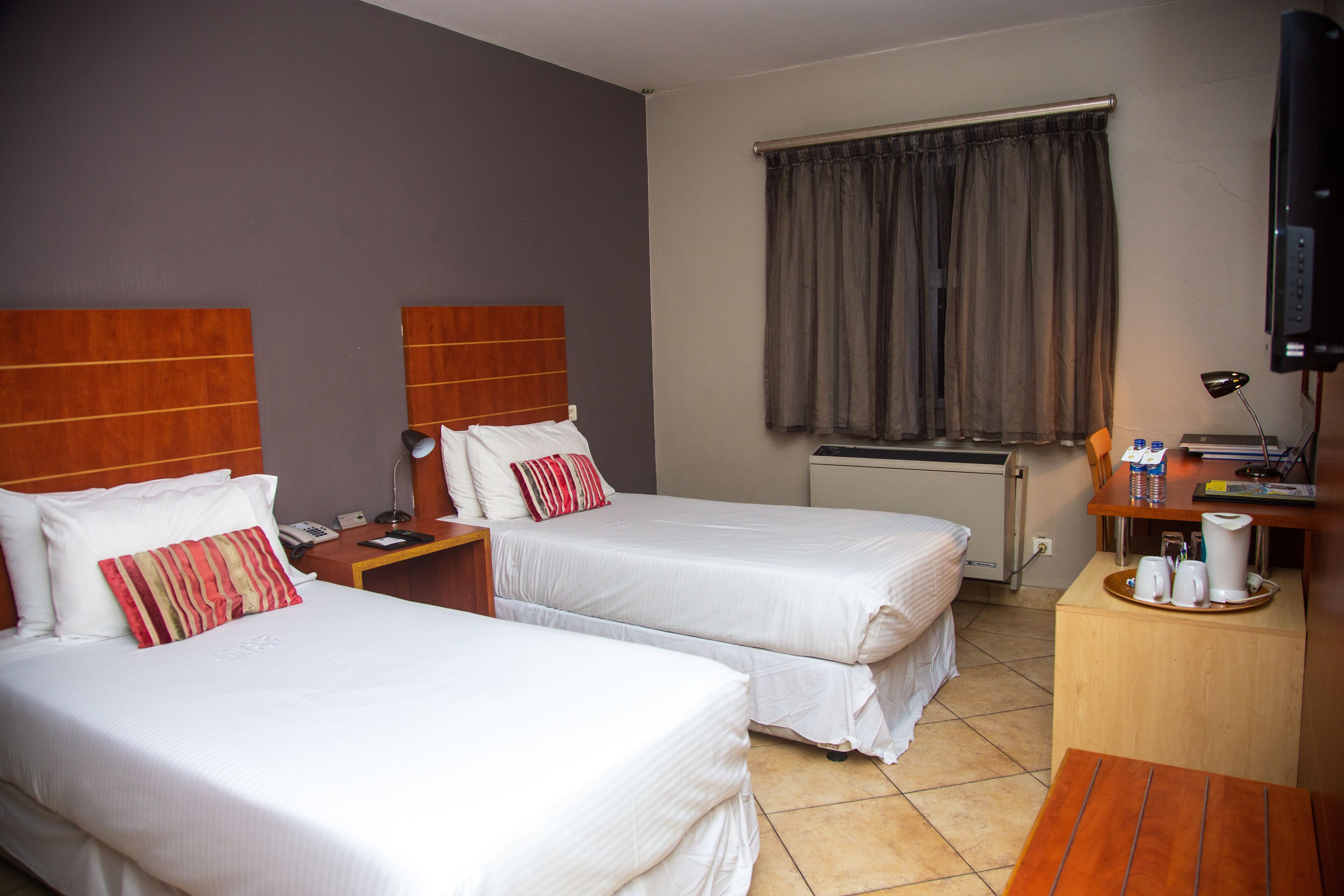 Quarto de hotel com duas camas, uma mesinha de centro com cafeteira, televisão e janela com cortinas.