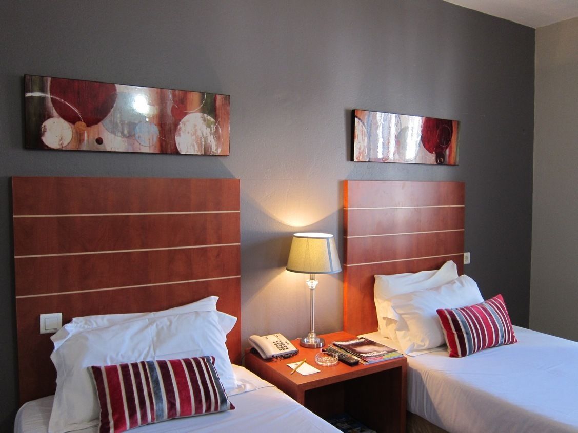 Quarto de hotel com duas camas, cabeceiras de madeira, mesa de cabeceira com abajur e obras de arte nas paredes.
