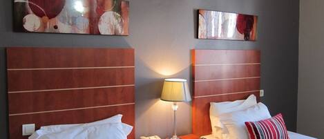Quarto de hotel com duas camas, cabeceiras de madeira, mesa de cabeceira com abajur e obras de arte nas paredes.