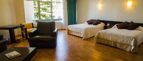 Quarto de hotel com duas camas, uma cadeira, uma mesa e uma janela com cortinas.