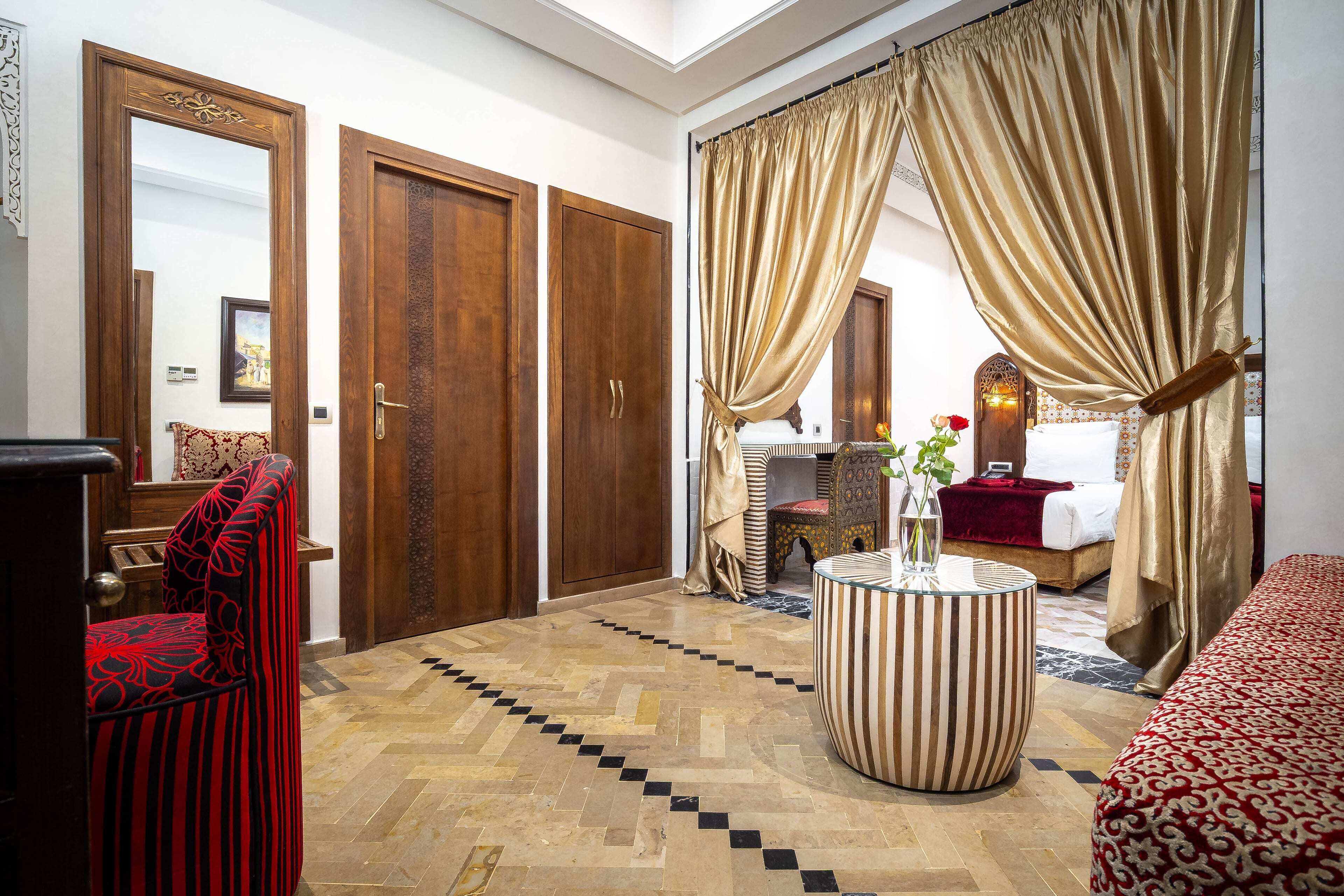 Quarto de hotel com padrão de piso em losango, mesa lateral redonda listrada, cama com colcha de padrão vermelho e branco e espelho grande com moldura de madeira.
