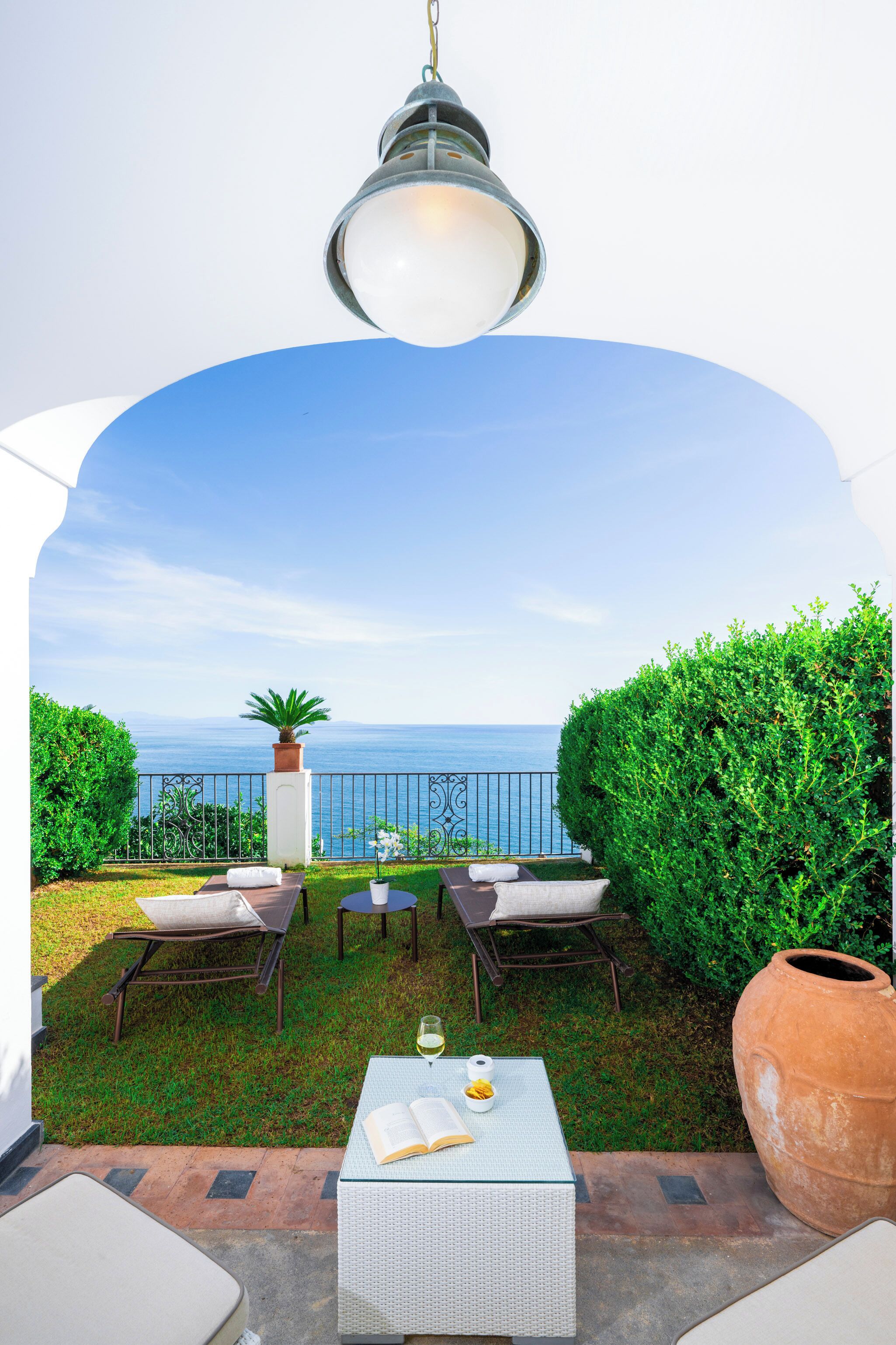 Um pátio com vista para o mar, mobiliado com cadeiras e uma mesa, e um candeeiro suspenso.