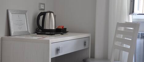 Quarto pequeno e bem arrumado, com uma escrivaninha, uma cadeira, um certificado emoldurado na parede e uma janela com cortinas.