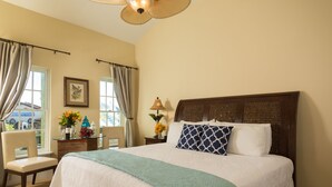 Quarto de hotel com uma cama grande, uma cadeira, escrivaninha com luminária e janela com cortinas.