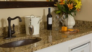 Uma bancada de cozinha com superfície em granito, uma pia branca, uma torneira estilo vintage, uma panela esmaltada branca, uma garrafa de vinho, dois copos de vinho e um vaso com flores.