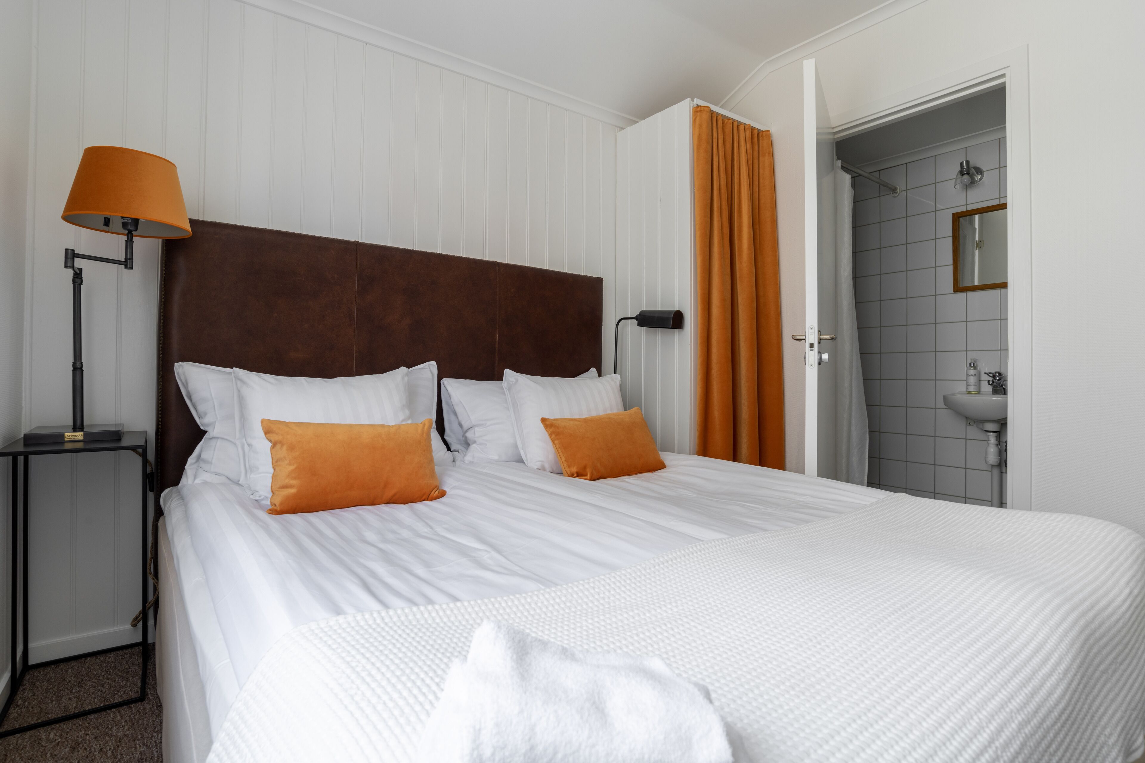 Cama bem arrumada com roupa de cama branca e laranja, abajur e banheiro visível através de uma porta aberta.