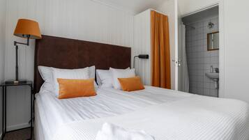 Cama bem arrumada com roupa de cama branca e laranja, abajur e banheiro visível através de uma porta aberta.