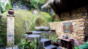 Um jardim com paredes de pedra, uma construção com telhado de palha, um espelho d'água e degraus que levam a uma área de estar.