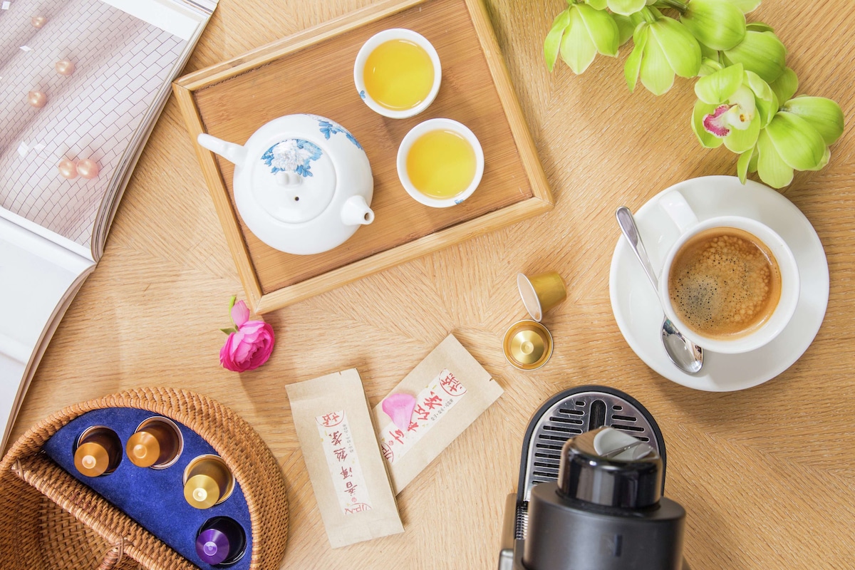 Uma bandeja de madeira com um bule, duas xícaras de chá, uma xícara de café, uma cafeteira, um livro, uma flor rosa e uma pequena cesta com velas.