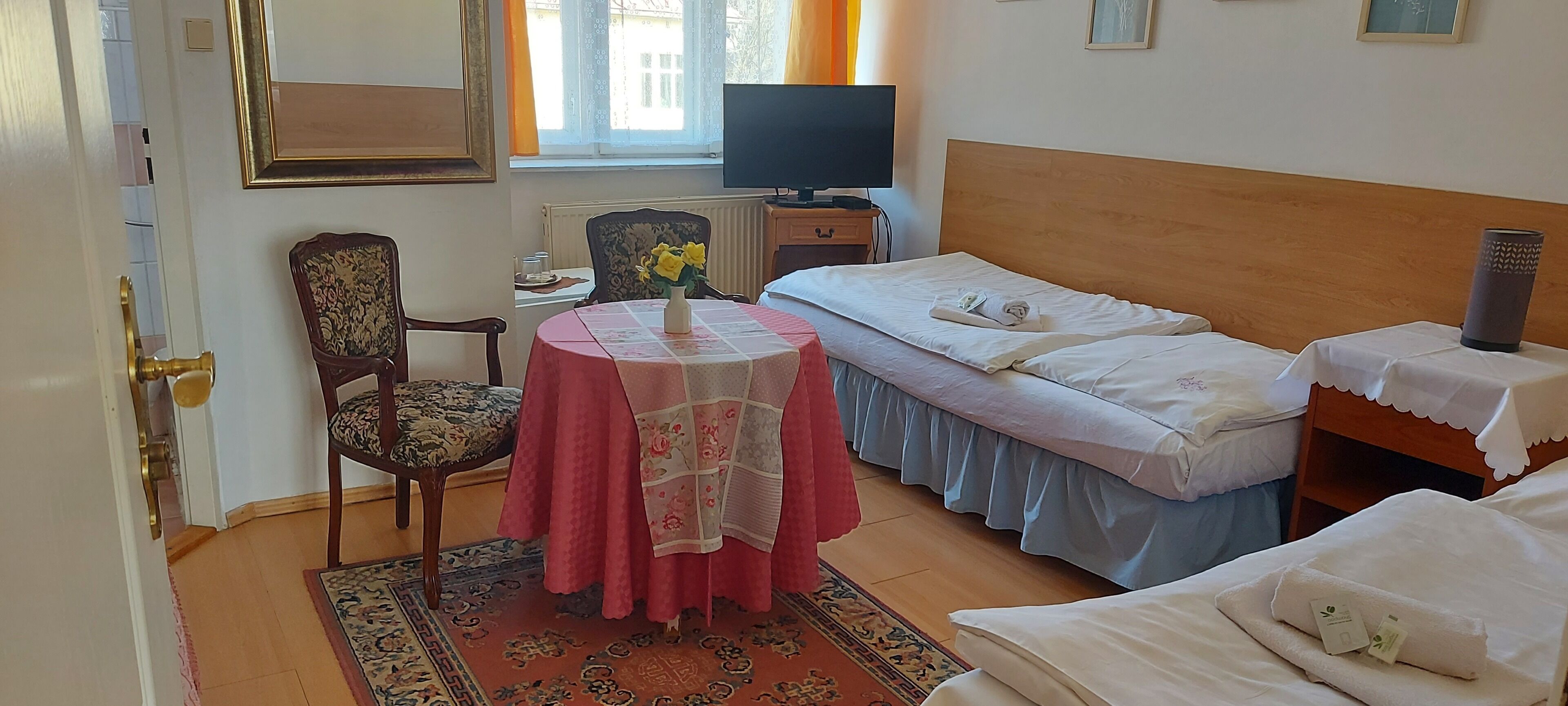 Quarto de hotel com cama, uma pequena mesa com um vaso de flores, uma cadeira, televisão e janela com cortinas.
