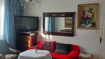 Uma sala de estar com televisão, sofá, poltrona, uma pequena mesa com um vaso e um quadro na parede.