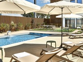 Piscina com espreguiçadeiras e guarda-sóis, cercada por um muro de madeira e vegetação.