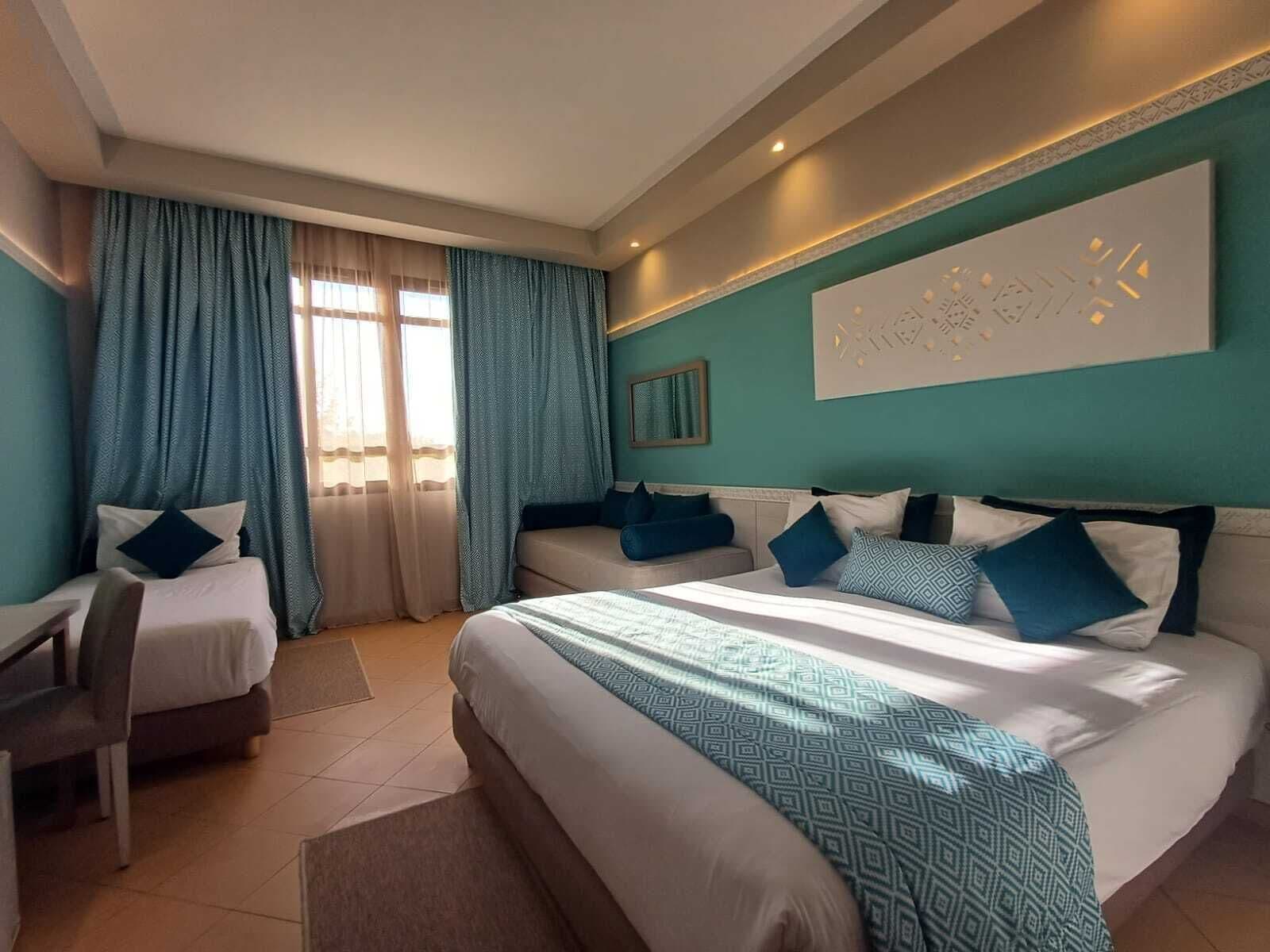 Quarto de hotel com duas camas, uma escrivaninha e uma janela grande com cortinas.