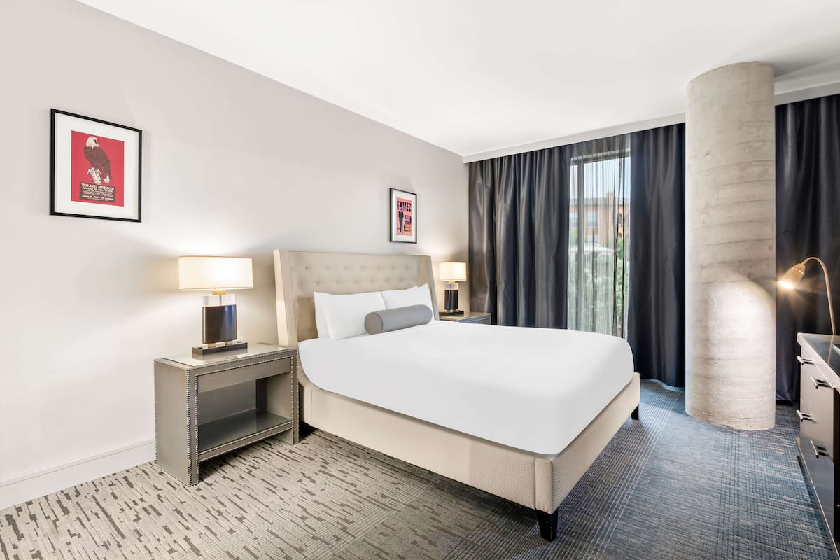 Quarto de hotel com uma cama grande, duas mesas de cabeceira com luminárias, uma janela ampla com cortinas e um pilar de concreto.