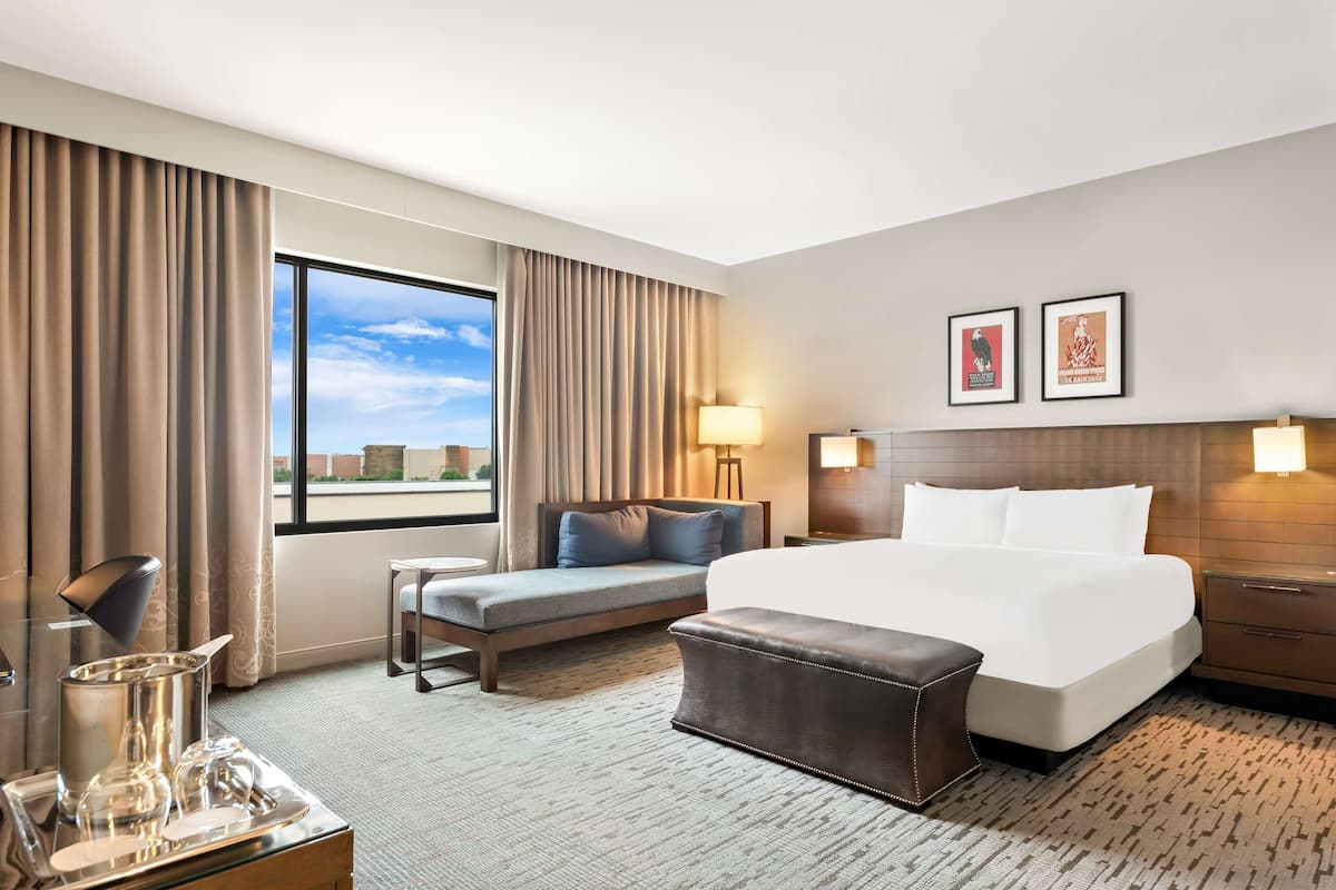 Quarto de hotel com uma cama grande, um sofá, uma escrivaninha com abajur e vista para a cidade.