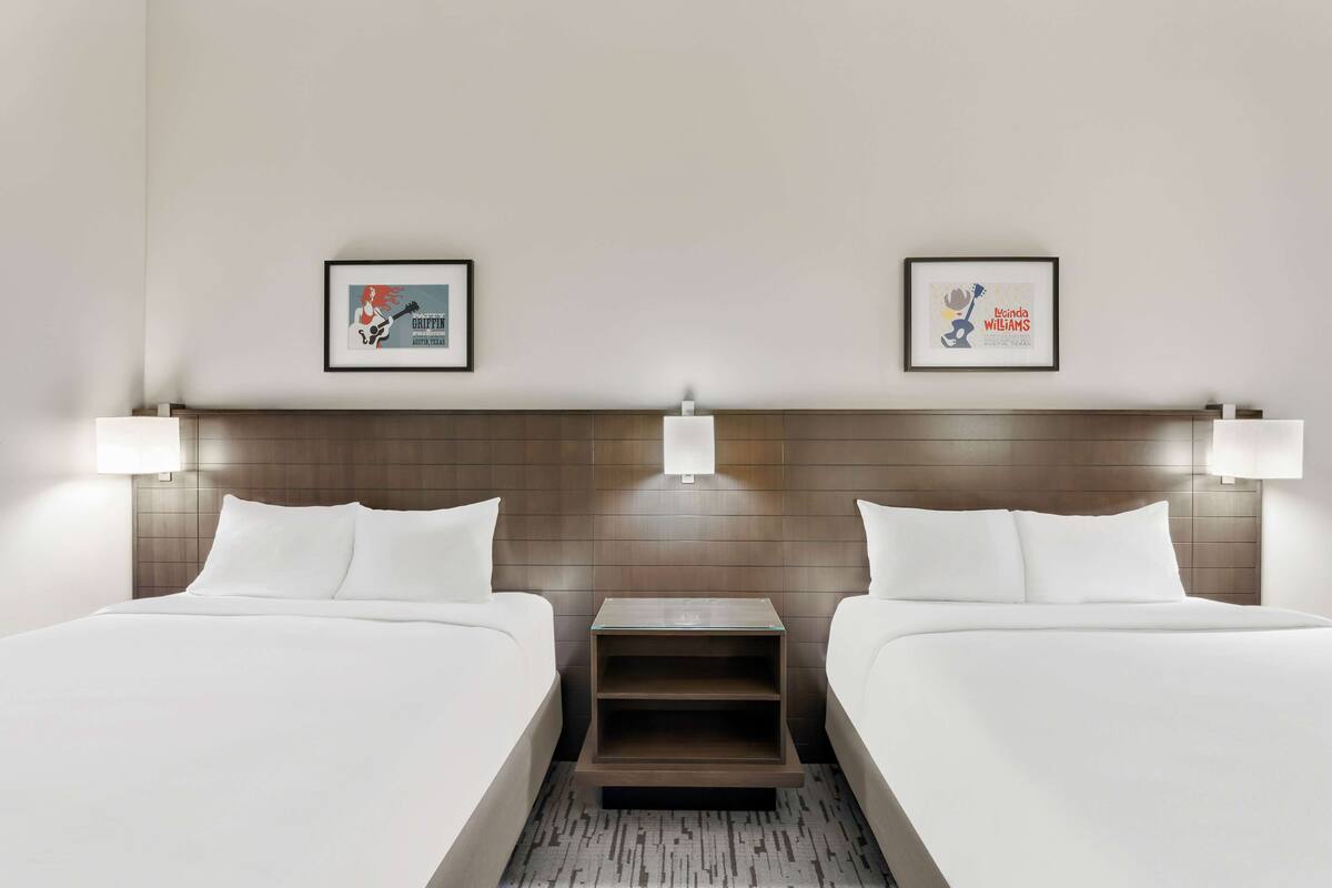 Quarto de hotel com duas camas, uma mesinha de cabeceira e luminárias fixadas na parede.