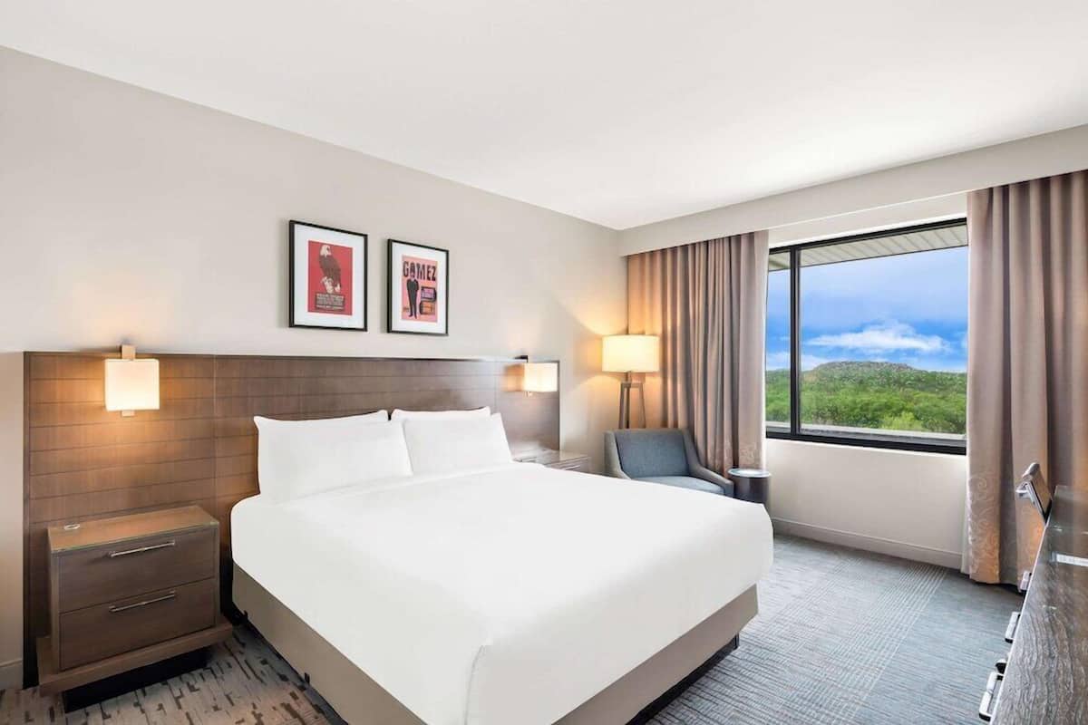 Quarto de hotel com uma cama grande, duas luminárias fixadas na parede, uma poltrona, mesa de cabeceira e uma janela ampla com vista.