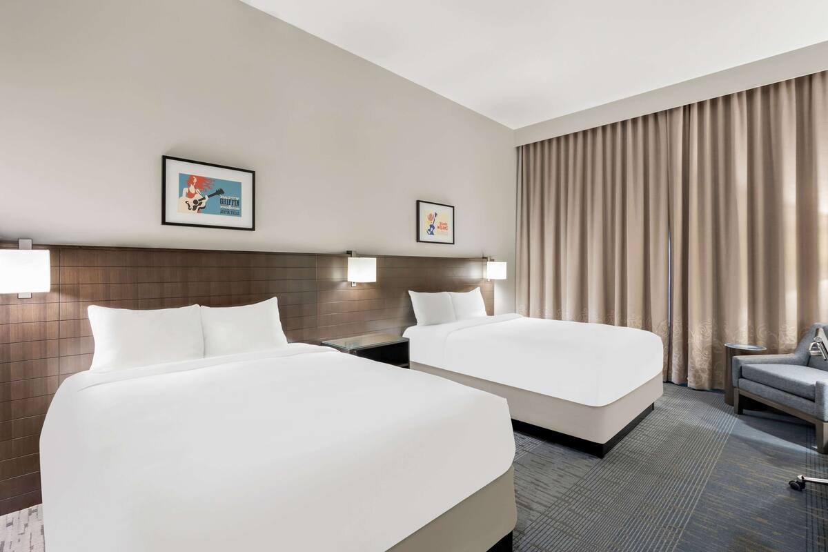 Quarto de hotel com duas camas, uma escrivaninha, uma cadeira e duas luminárias fixadas na parede.