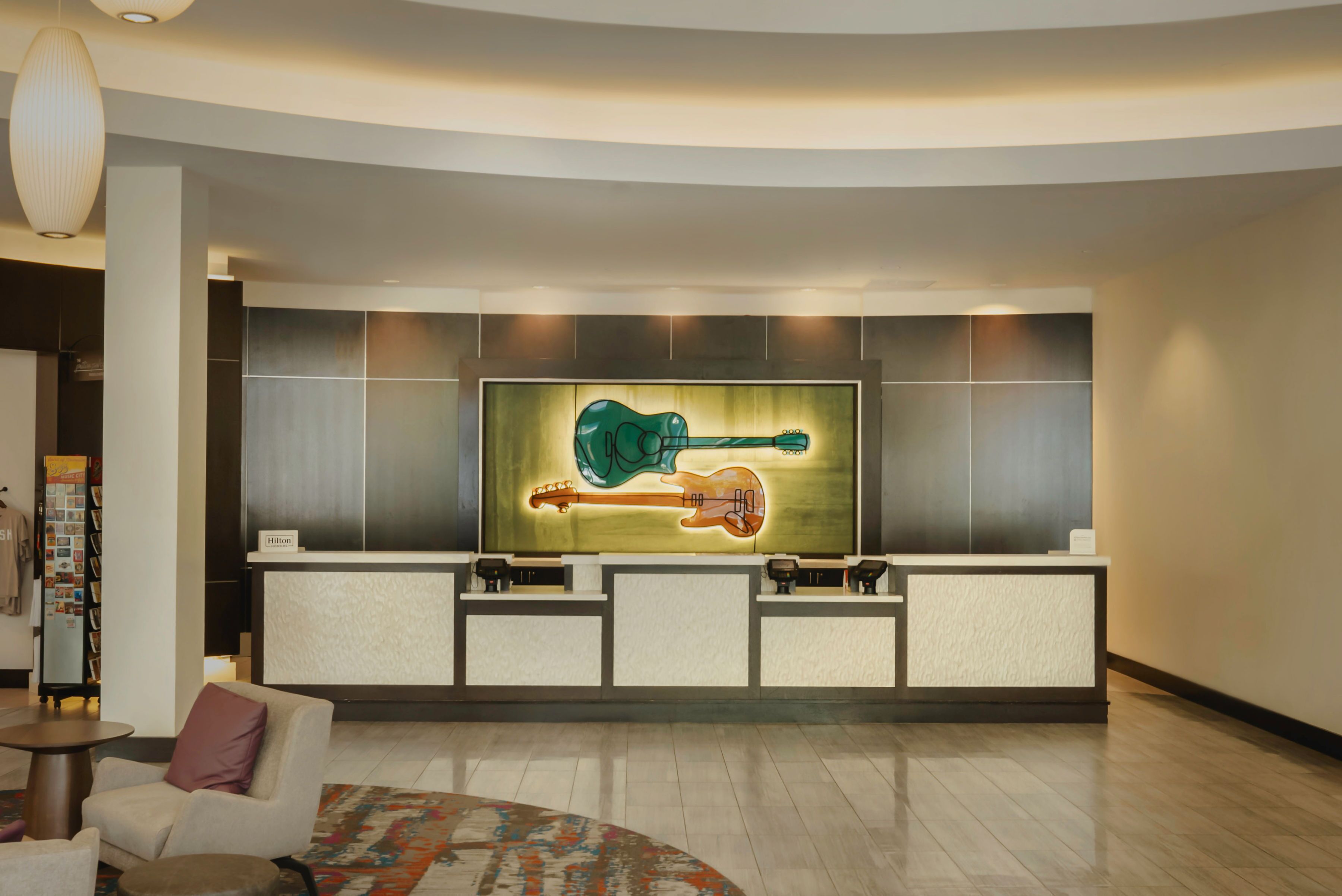 Uma área de recepção de hotel com uma grande obra de arte fixada na parede, representando uma guitarra, um balcão de recepção e uma área de estar com uma mesa redonda e cadeiras.