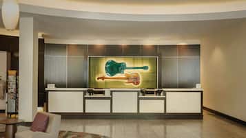 Uma área de recepção de hotel com uma grande obra de arte fixada na parede, representando uma guitarra, um balcão de recepção e uma área de estar com uma mesa redonda e cadeiras.