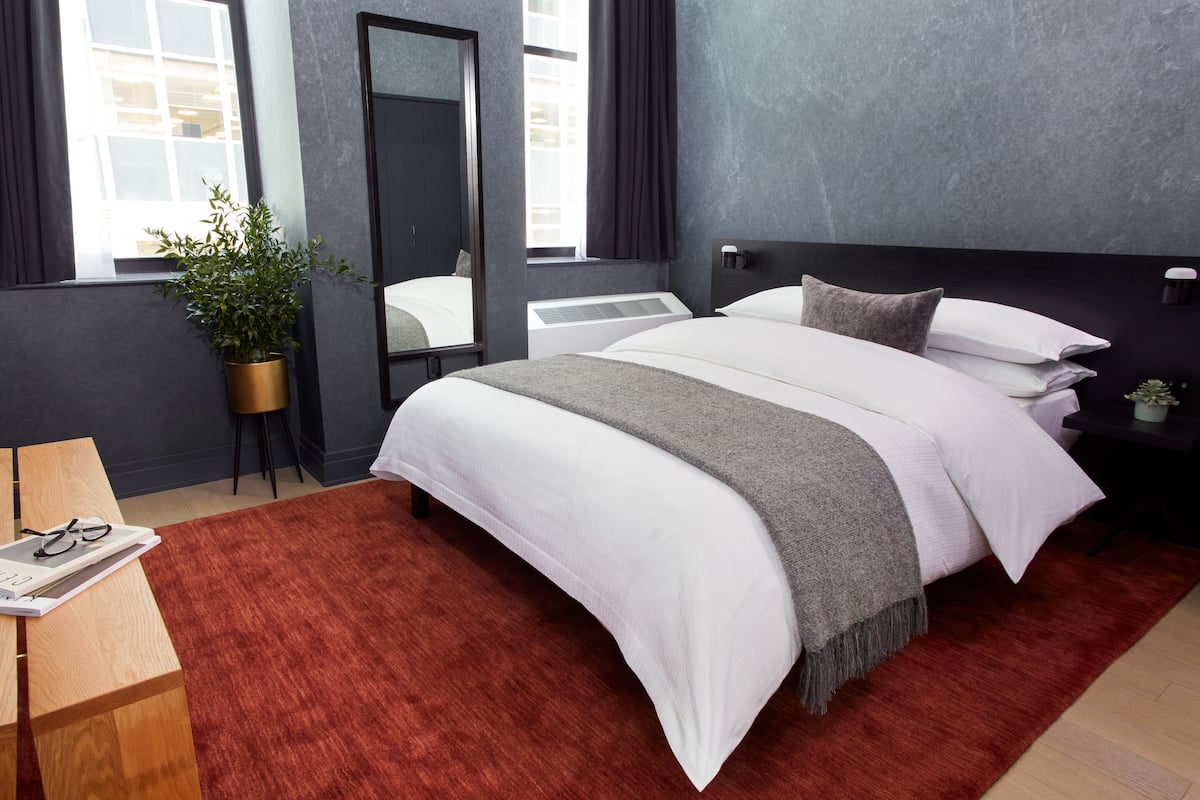 Quarto de hotel moderno com uma cama grande, uma mesa, uma cadeira, uma planta em vaso e vista para a cidade.