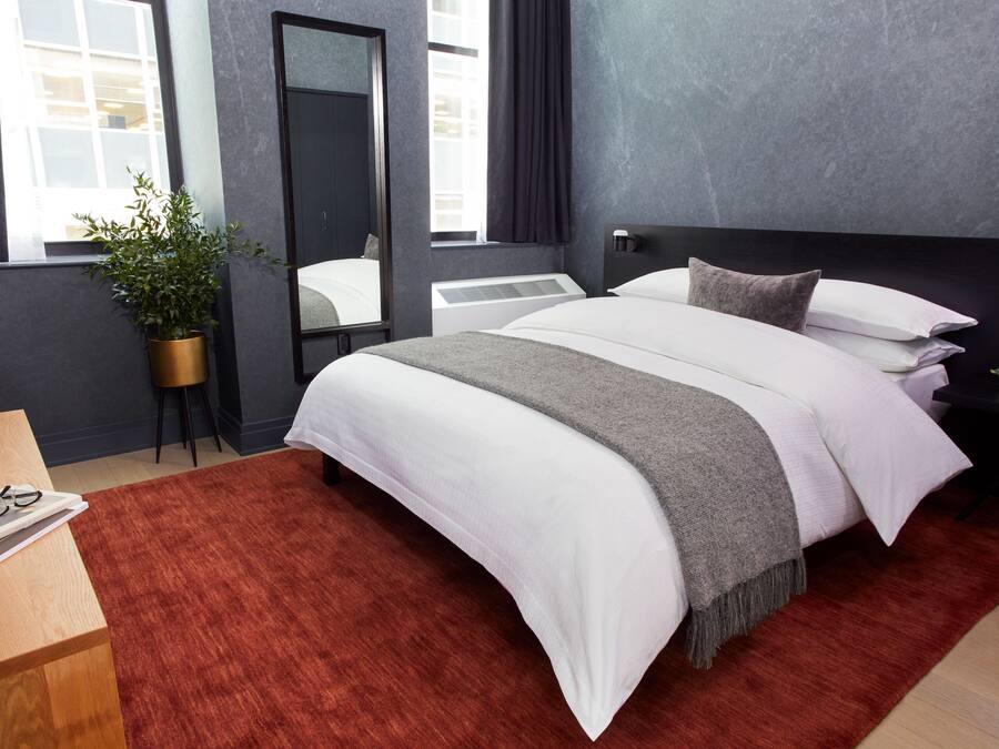 Quarto de hotel moderno com uma cama grande, uma mesa, uma cadeira, uma planta em vaso e vista para a cidade.