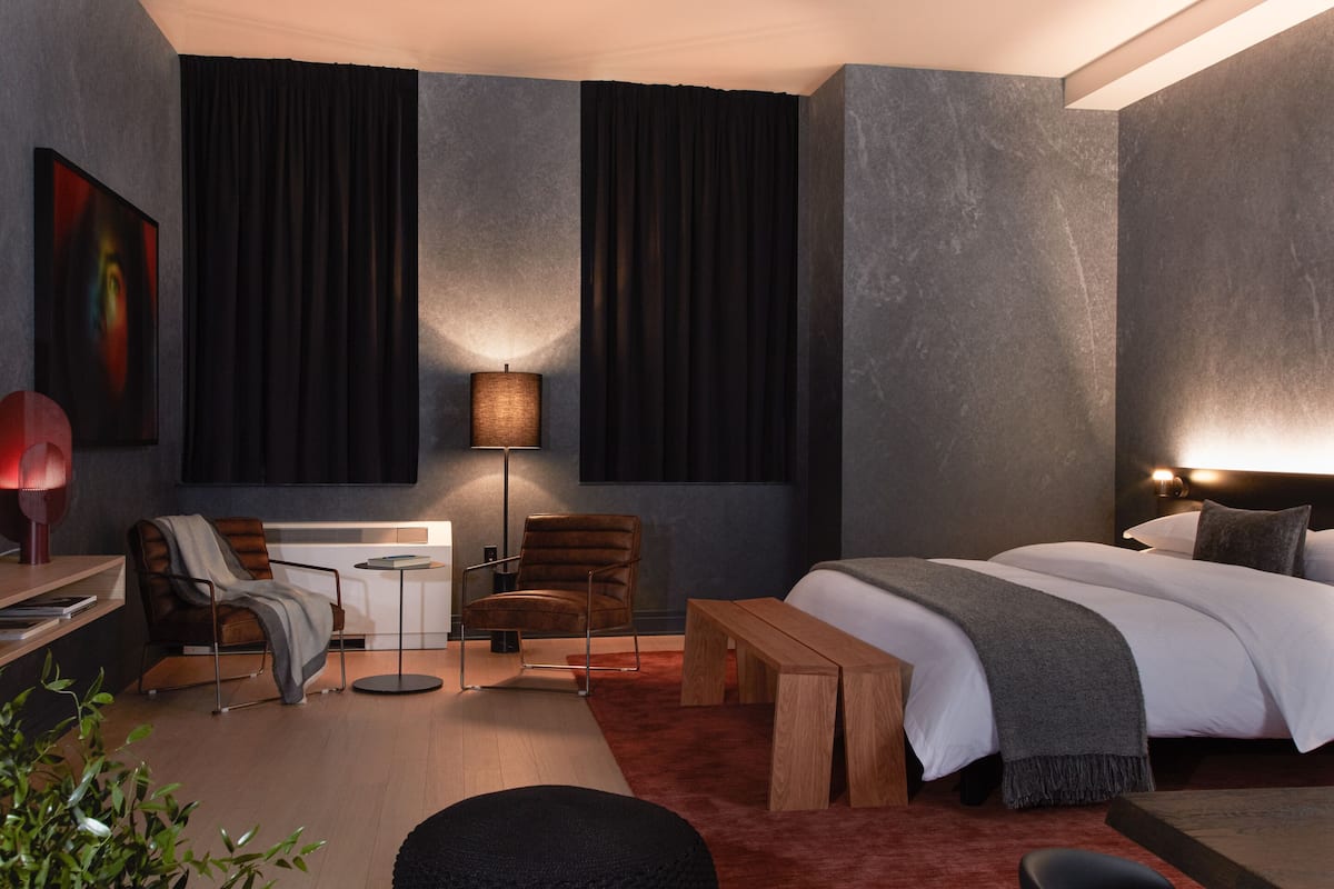 Quarto de hotel moderno com cama, escrivaninha, cadeiras e luminária.