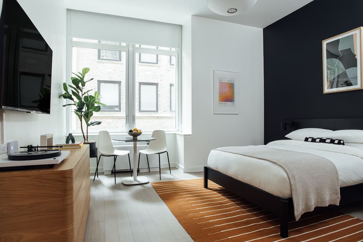 Quarto moderno com cama, escrivaninha com computador, cadeira e janela com vista para edifícios.