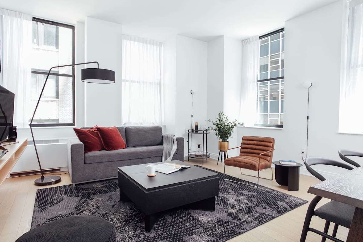 Uma sala de estar moderna com um sofá cinza, mesa de centro preta e uma mesa de jantar. Há duas cadeiras, um abajur e um vaso de planta.