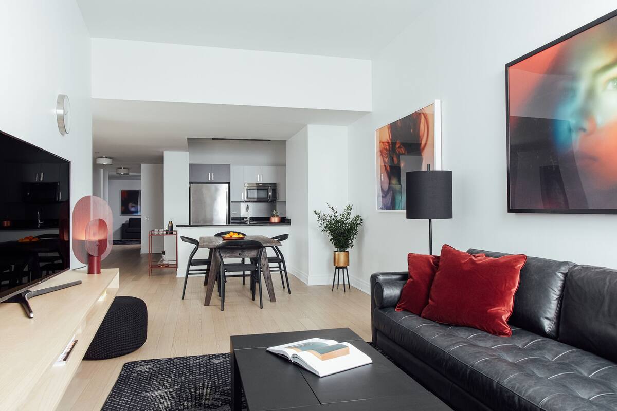 Uma sala de estar moderna com um sofá de couro preto, uma mesa de jantar com cadeiras, uma cozinha ao fundo e um grande quadro abstrato na parede.