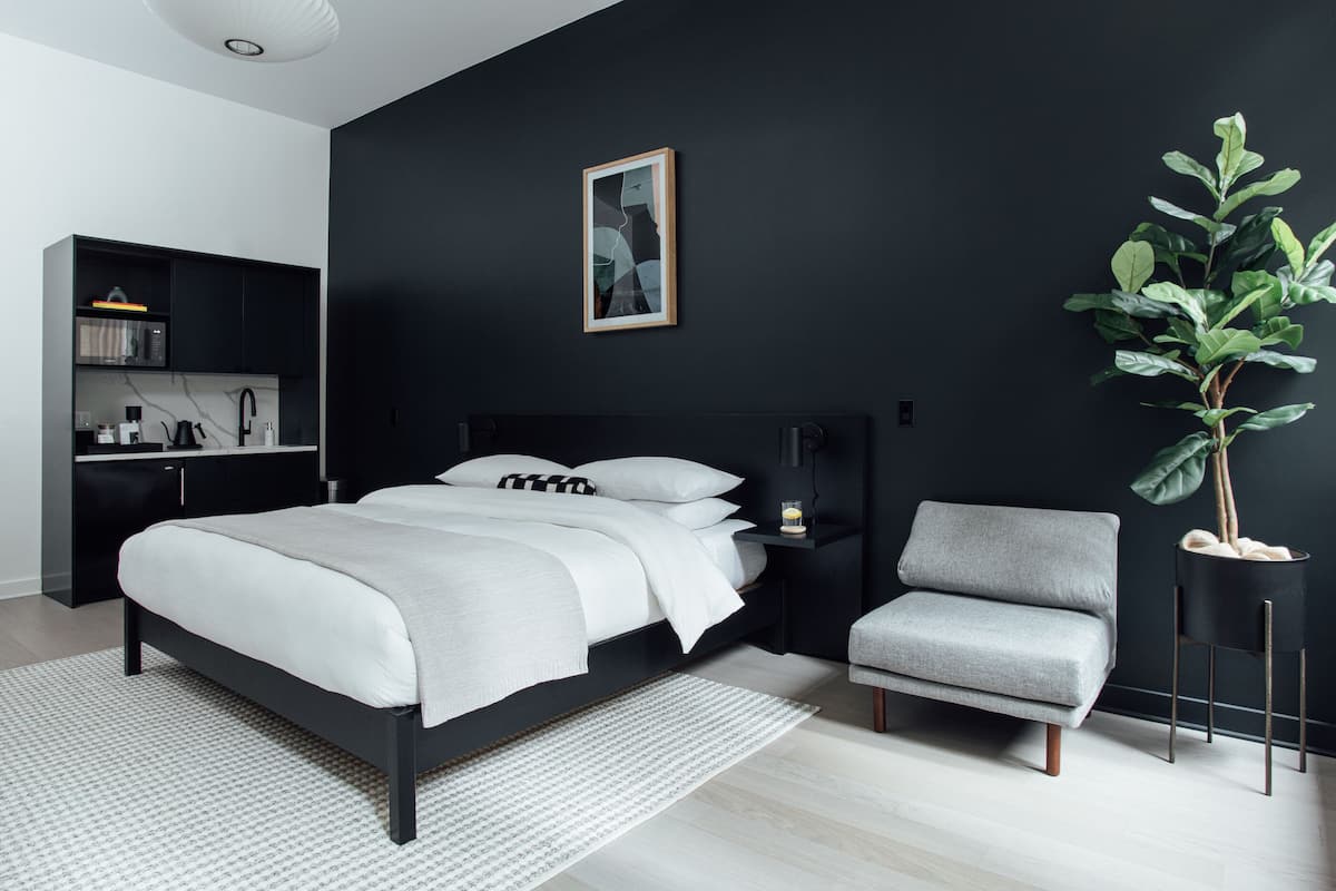 Quarto moderno com parede preta, cama com roupa de cama branca, poltrona cinza, mesa de cabeceira com abajur e uma planta.