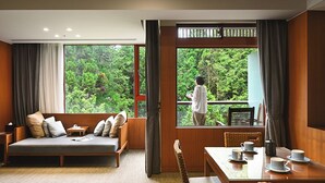 Quarto com uma janela grande, um sofá, uma mesa de jantar com cadeiras e vista para o verde lá fora.