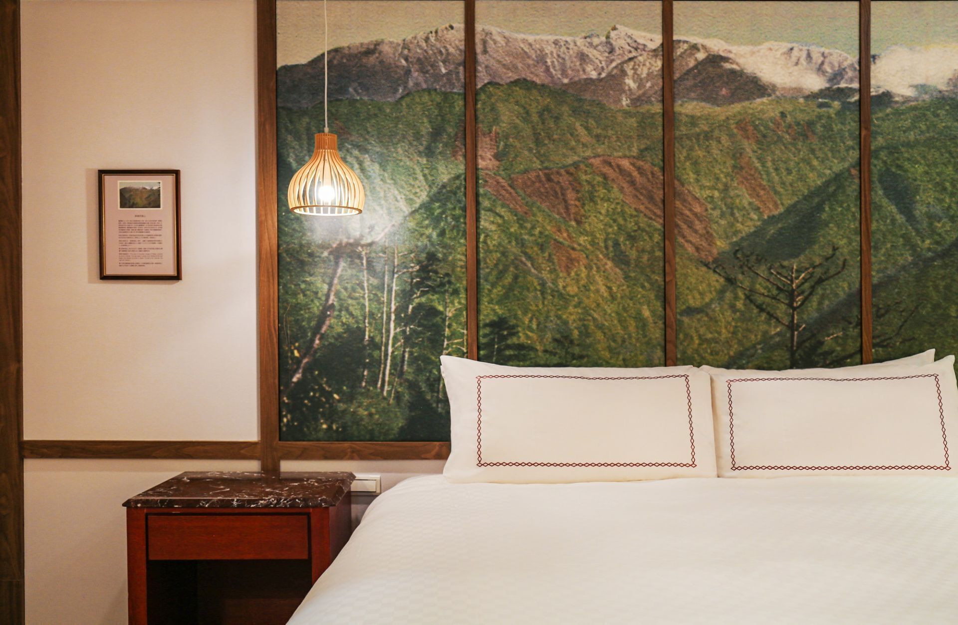 Quarto de hotel com cama, mesa de cabeceira, quadro pendurado na parede e luminária.