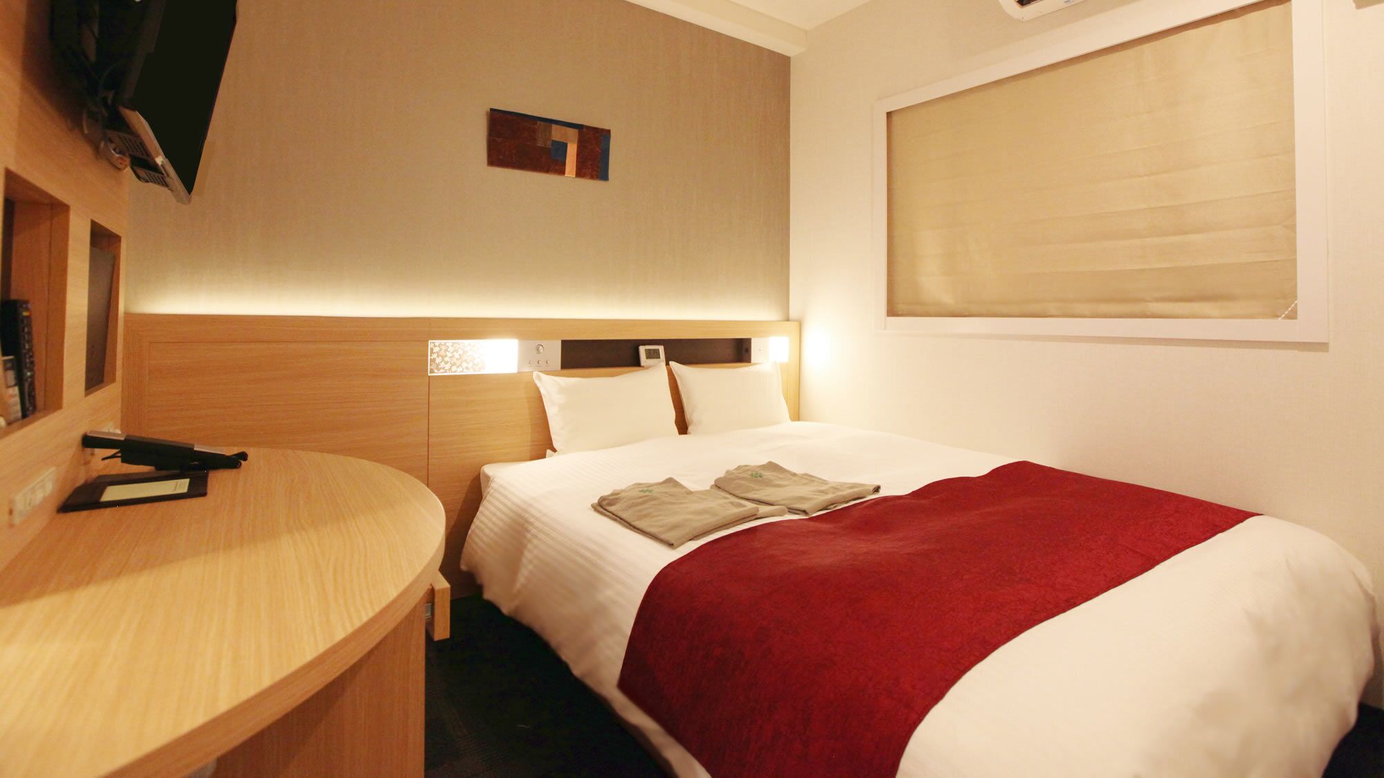 Quarto de hotel com cama, escrivaninha, televisão e janela com persianas.