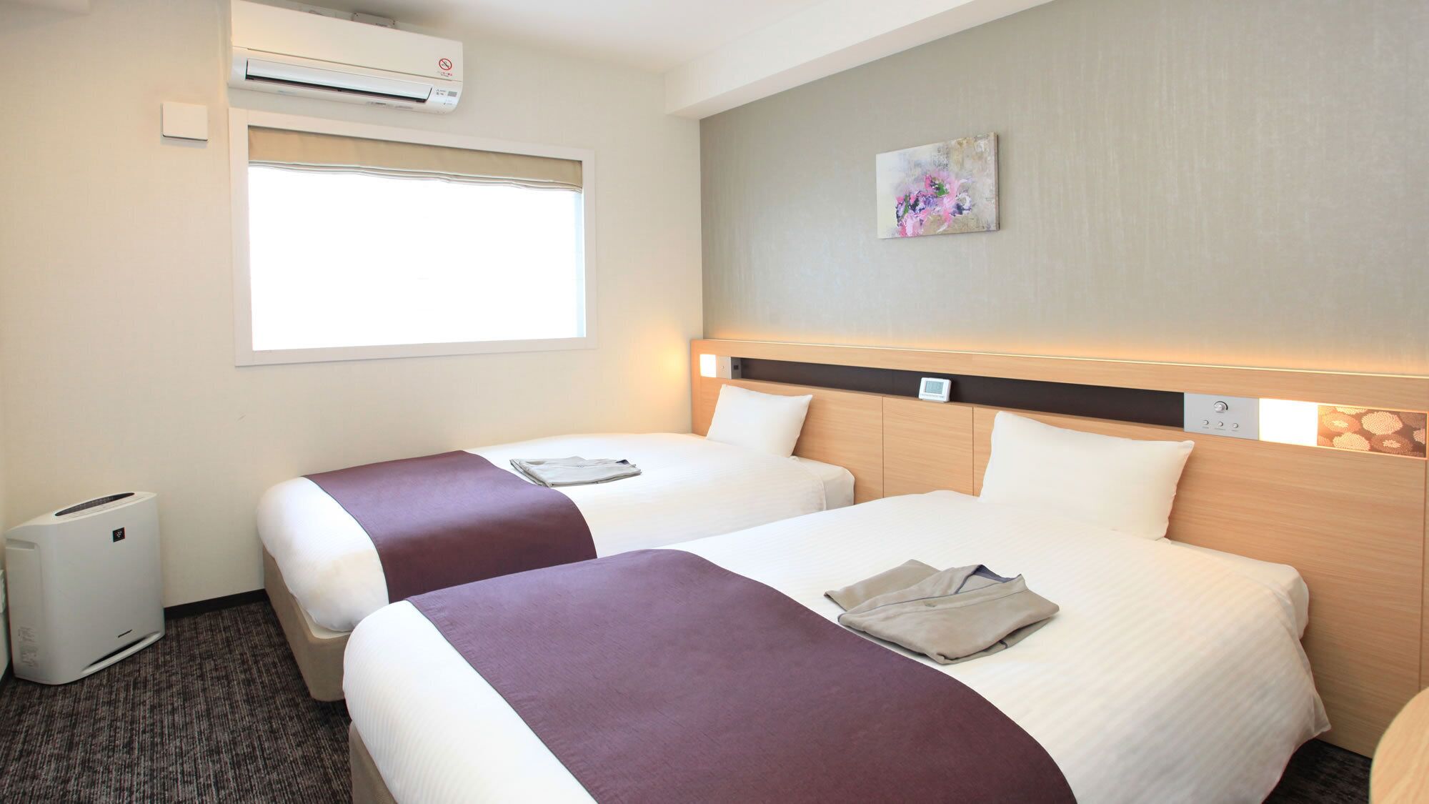Quarto de hotel com duas camas, uma janela, ar condicionado e um quadro na parede.