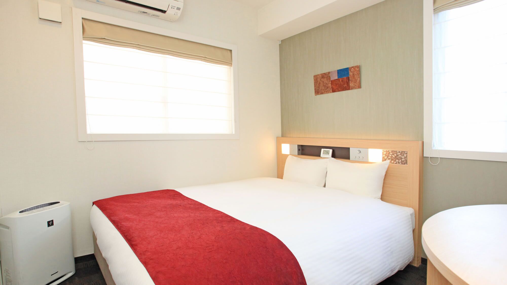 Quarto de hotel com cama, mesa de cabeceira, janela com persianas, aparelho de ar condicionado e uma obra de arte na parede.