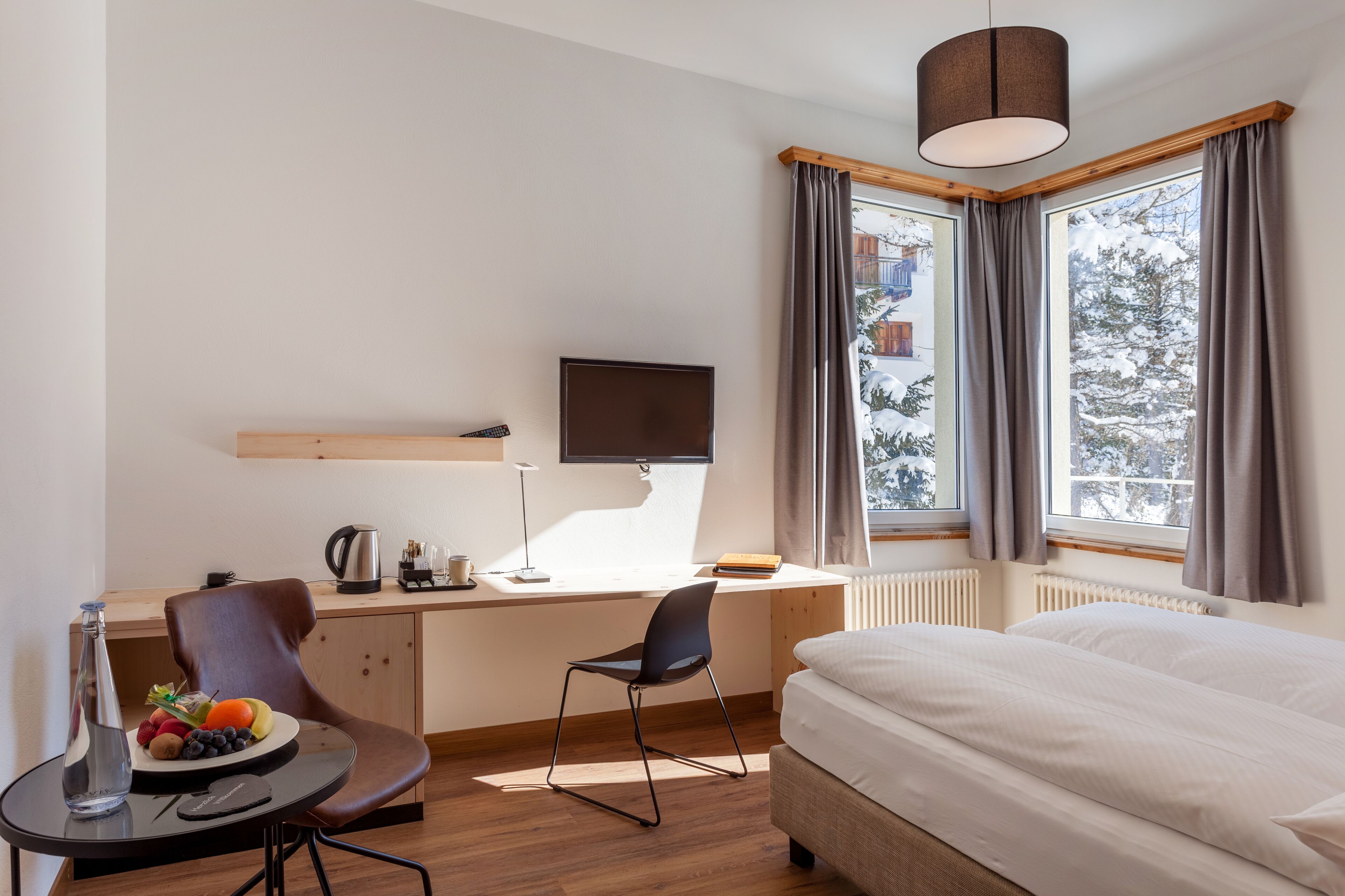 Quarto de hotel com cama, escrivaninha, cadeira, televisão e janela com cortinas.