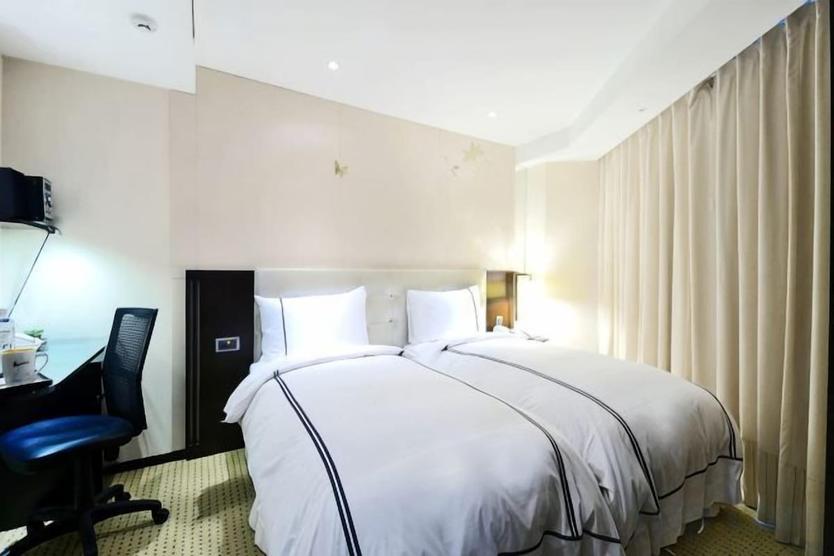 Quarto de hotel com cama de casal, escrivaninha com cadeira, luminária e janela com cortinas.