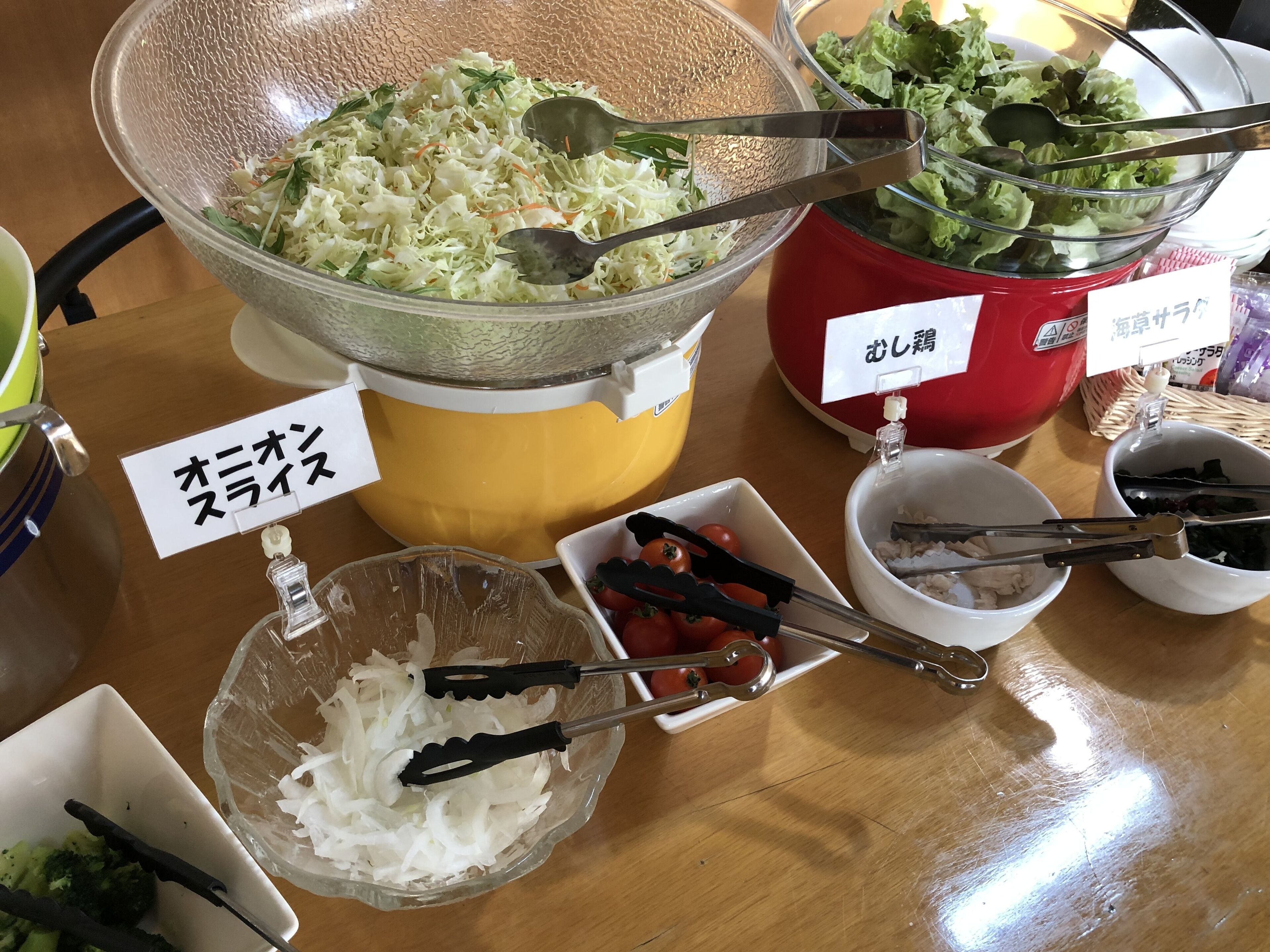 Uma mesa com diversos alimentos e utensílios, incluindo uma tigela grande de salada de repolho, uma salada de folhas e uma tigela de arroz.