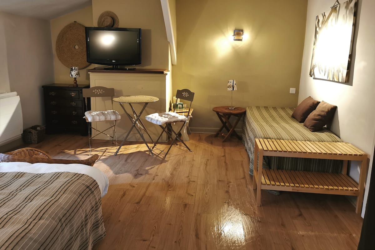 Quarto aconchegante com uma cama, uma mesa pequena com cadeiras, uma televisão e uma luminária na parede.