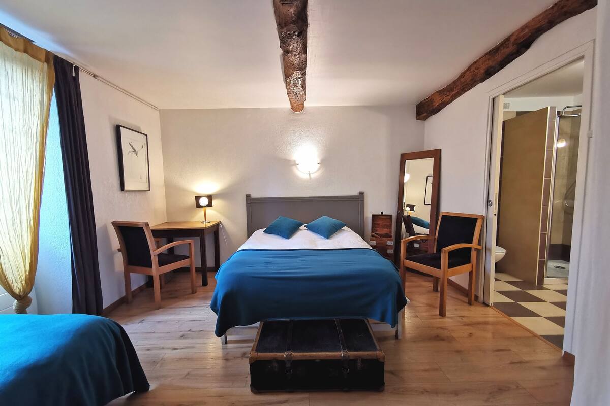 Quarto de hotel com cama, escrivaninha, cadeira, mala e área de chuveiro.