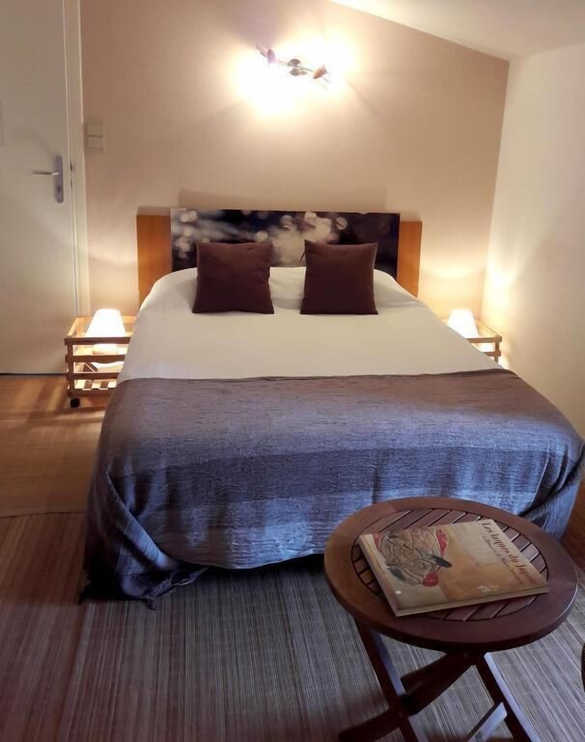 Cama bem arrumada com manta branca e cinza, duas almofadas castanhas e cabeceira com desenho floral. Duas luminárias de cabeceira estão sobre mesas de cabeceira de madeira. Uma pequena mesa redonda com uma revista está em frente à cama.