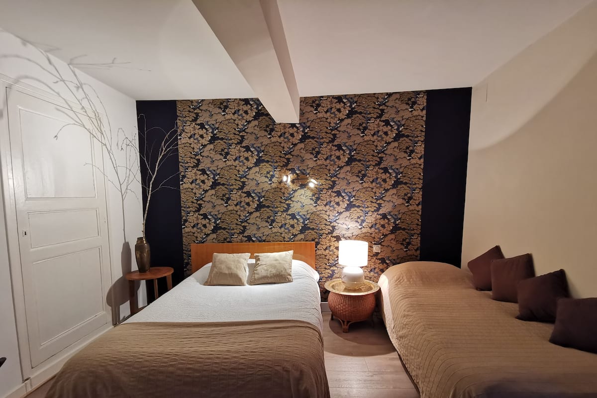 Quarto com duas camas, mesa de cabeceira com abajur, parede decorativa e pequena mesa com um vaso.
