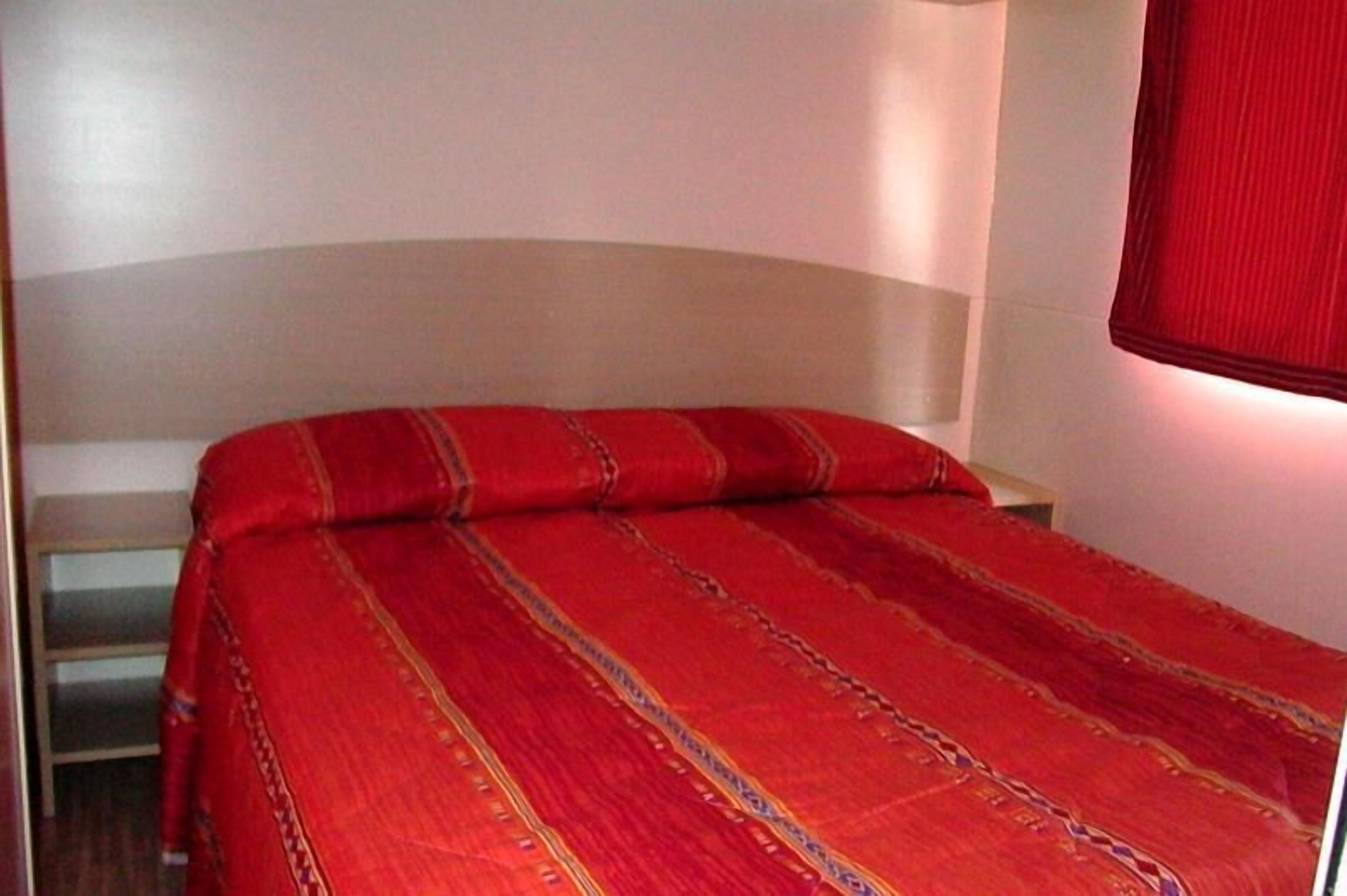 Uma cama com um cobertor vermelho num quarto com parede branca e cortina vermelha.