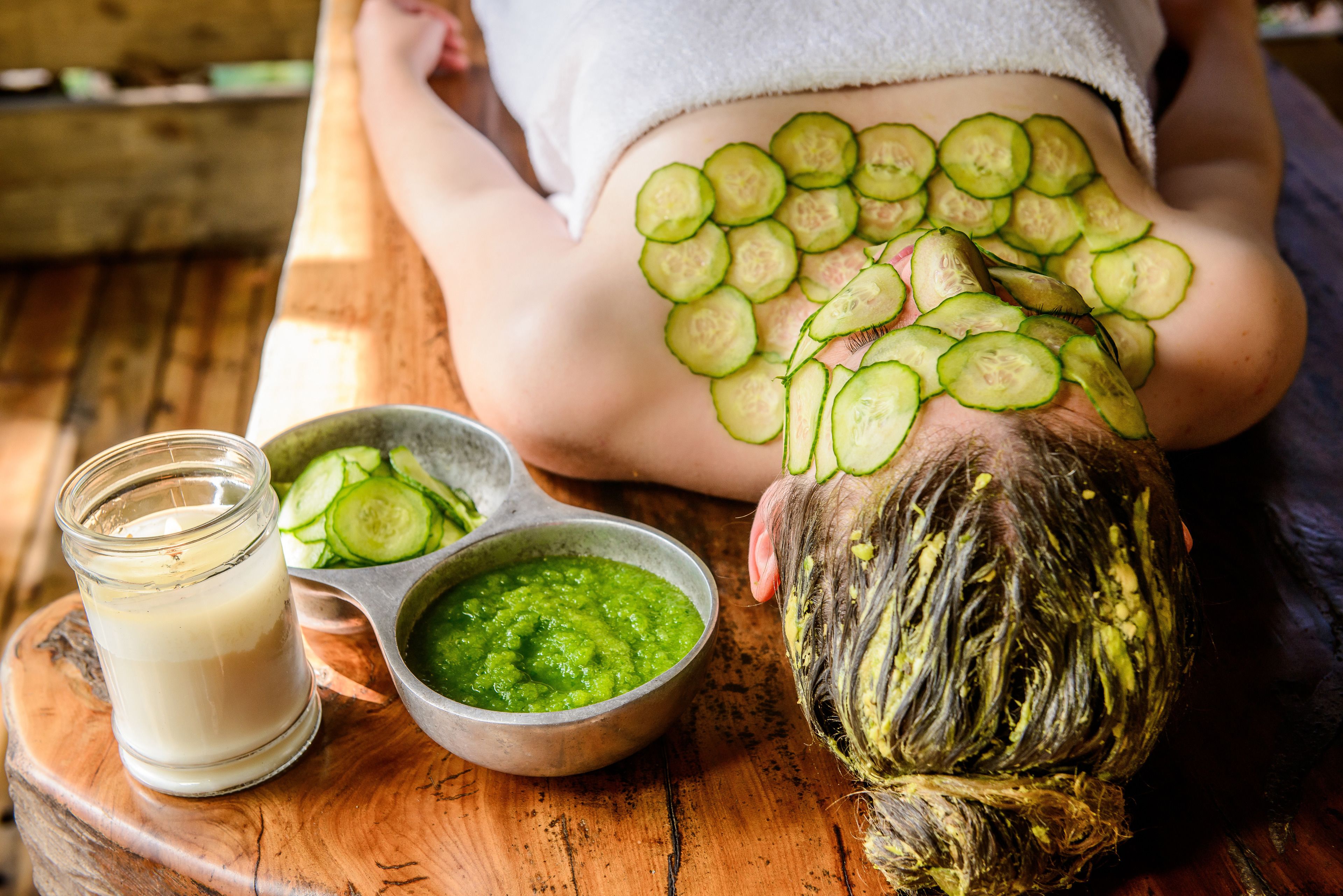 Uma pessoa recebendo um tratamento com pepino, com recipientes de pasta verde e um frasco de líquido sobre uma superfície de madeira.