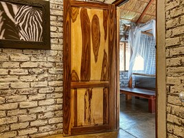 Uma porta de madeira rústica com um padrão de veios único, que dá acesso a um quarto com parede de pedra e uma cama com dossel.